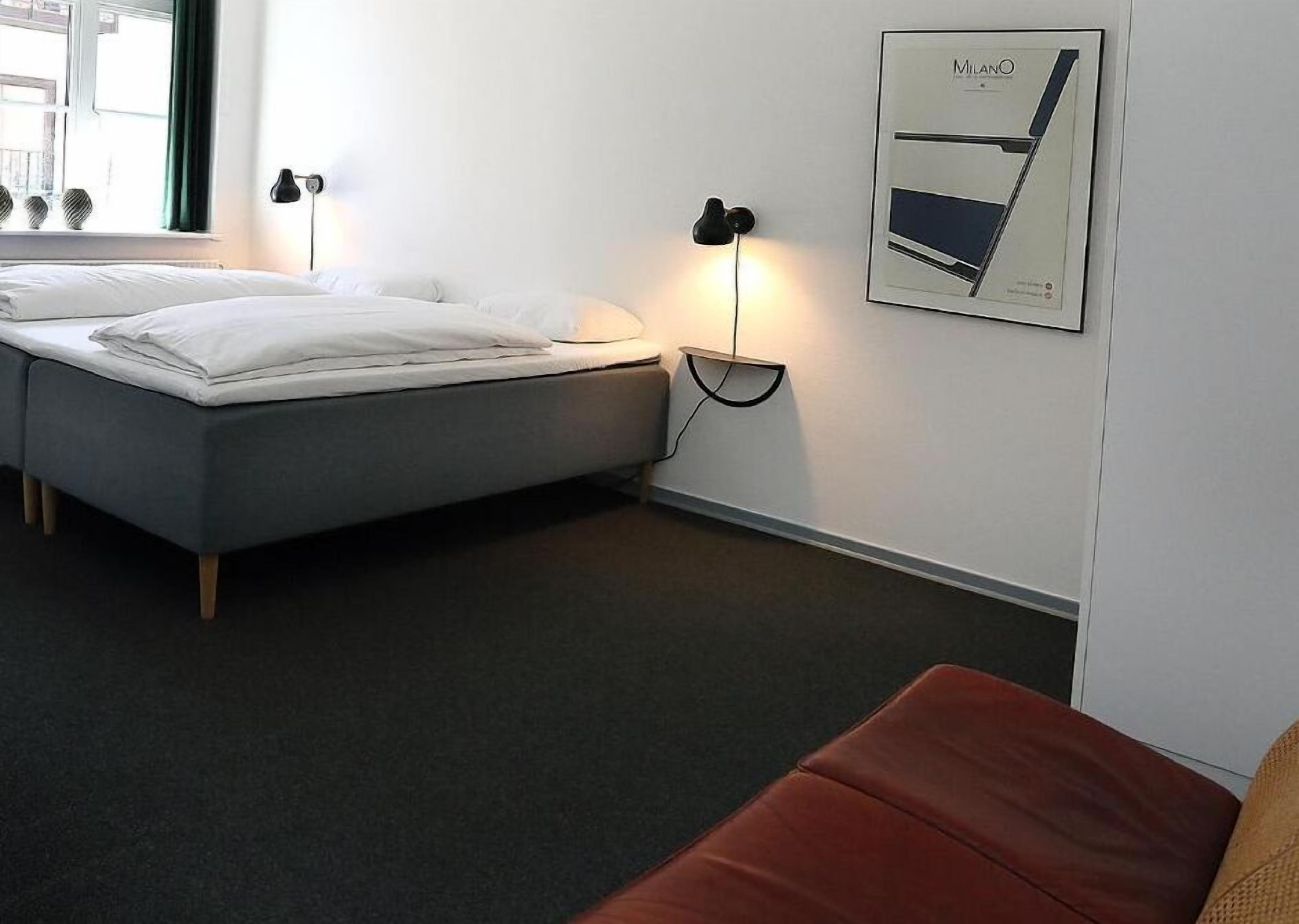 Quarto de hotel com cama, abajures, luz acoplada na parede e um quadro emoldurado.