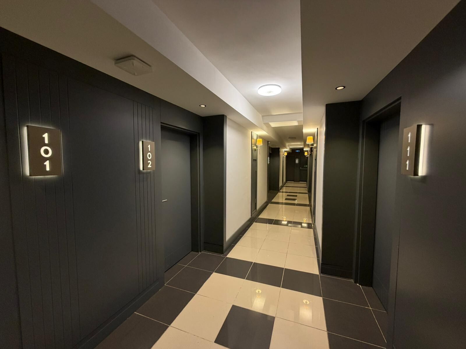 Um corredor de hotel moderno com piso xadrez preto e branco, paredes escuras e números de quarto iluminados.