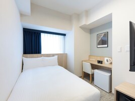 Quarto de hotel com cama, escrivaninha, cadeira, pequeno frigorífico e janela com cortinas.