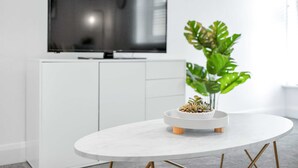Uma sala de estar moderna com uma TV de tela plana fixada em um móvel branco, uma mesa de mármore redonda com uma planta em vaso e uma grande janela com iluminação natural.