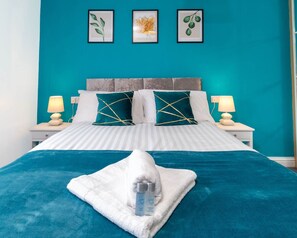 Quarto de hotel com parede verde-água, uma cama com roupa de cama listrada e duas mesas de cabeceira com abajures. Há três quadros na parede acima da cama.