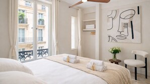 Quarto de hotel com cama, ventilador de teto, vista para o exterior, parede com arte minimalista e uma pequena mesa com um vaso.