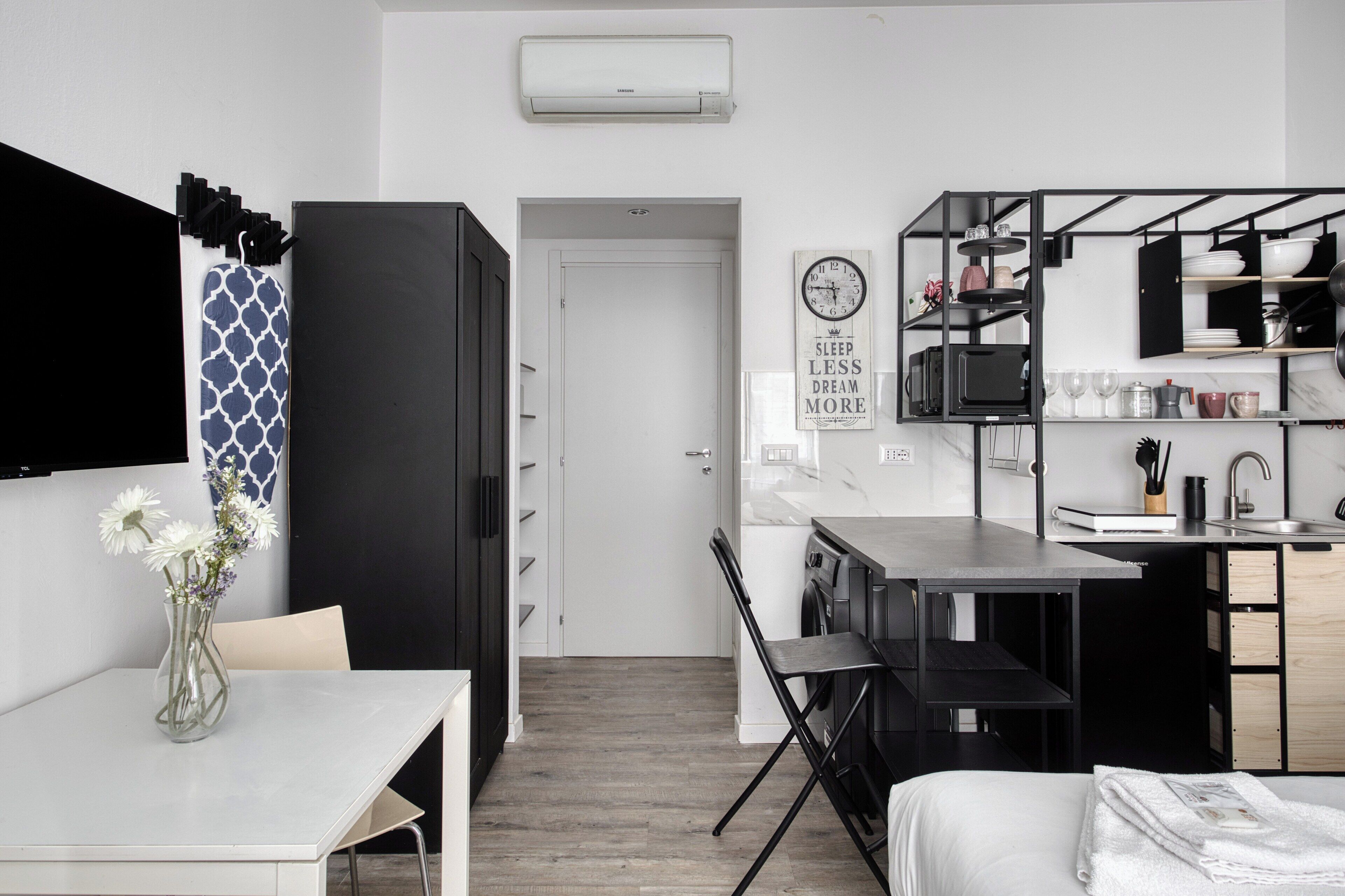 Uma kitchenette compacta com micro-ondas, pia e prateleiras de armazenamento. Há uma área de refeições com mesa e cadeiras, e um pequeno armário branco.