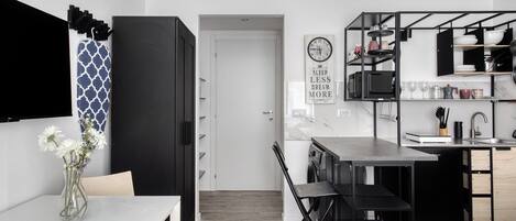 Uma kitchenette compacta com micro-ondas, pia e prateleiras de armazenamento. Há uma área de refeições com mesa e cadeiras, e um pequeno armário branco.