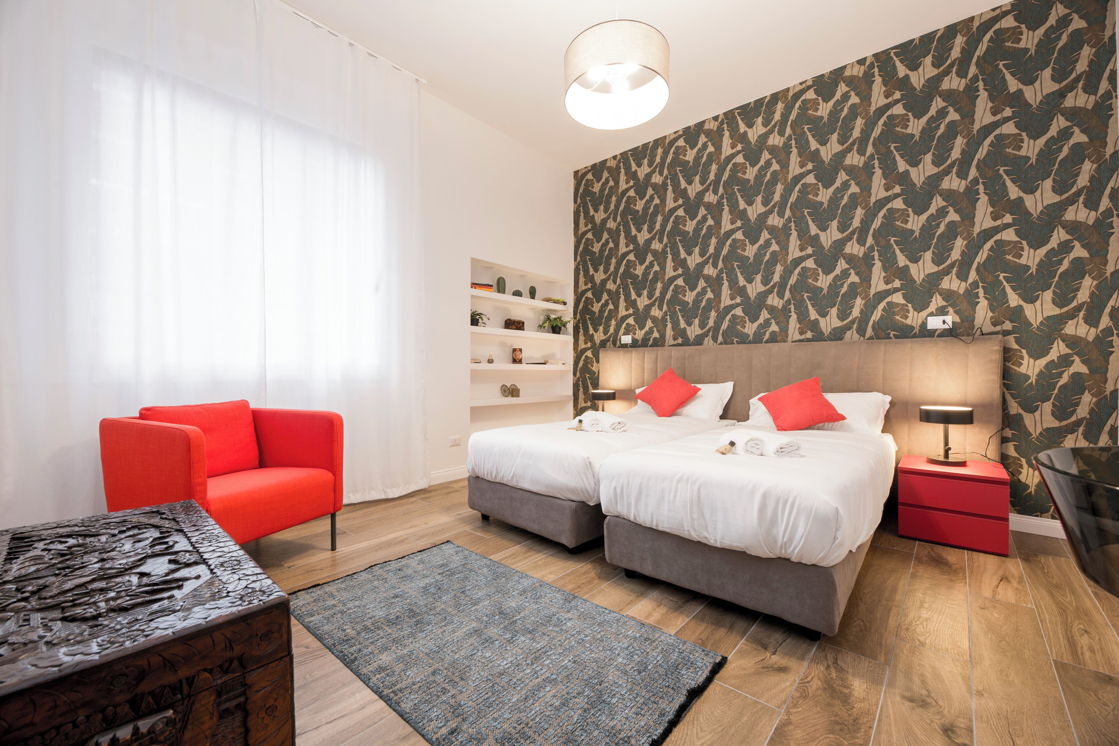 Quarto moderno com duas camas, um sofá vermelho, uma mesa de centro de madeira e uma parede estampada.