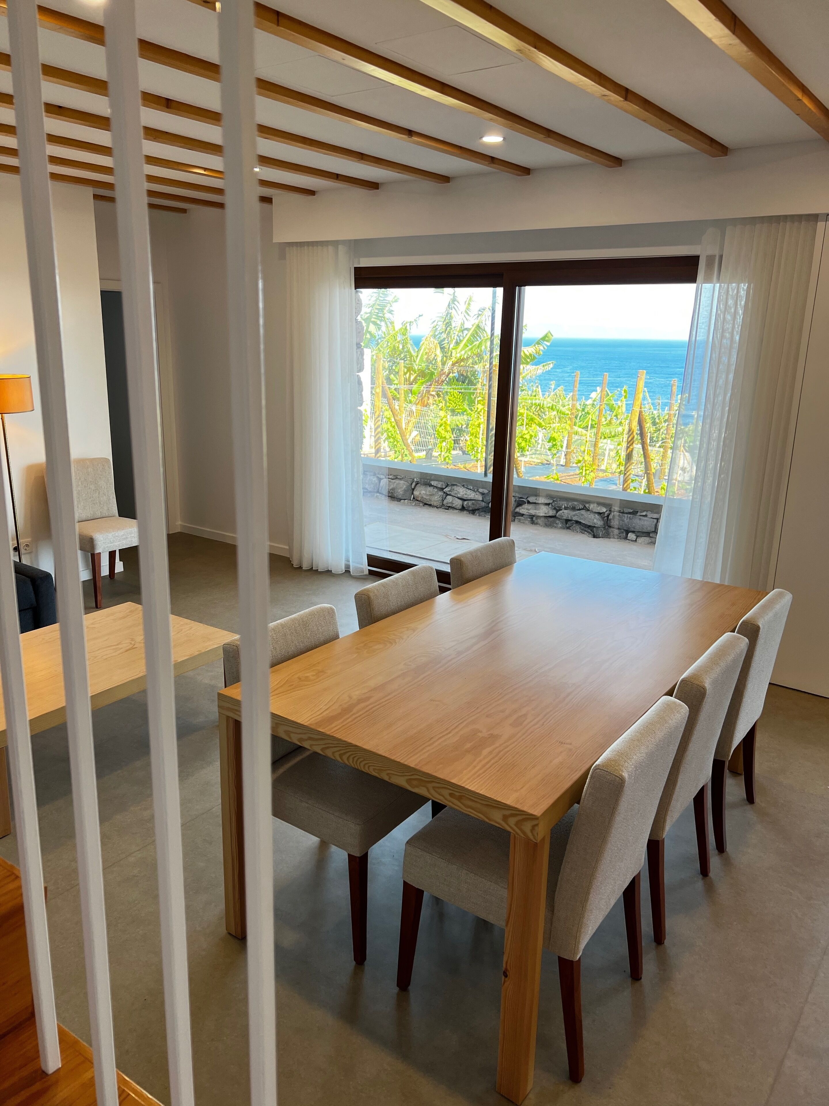 Uma sala de jantar moderna com mesa e cadeiras de madeira, vista para o oceano através de grandes janelas e uma parede de pedra ao fundo.