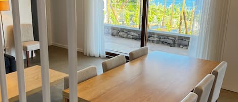 Uma sala de jantar moderna com mesa e cadeiras de madeira, vista para o oceano através de grandes janelas e uma parede de pedra ao fundo.