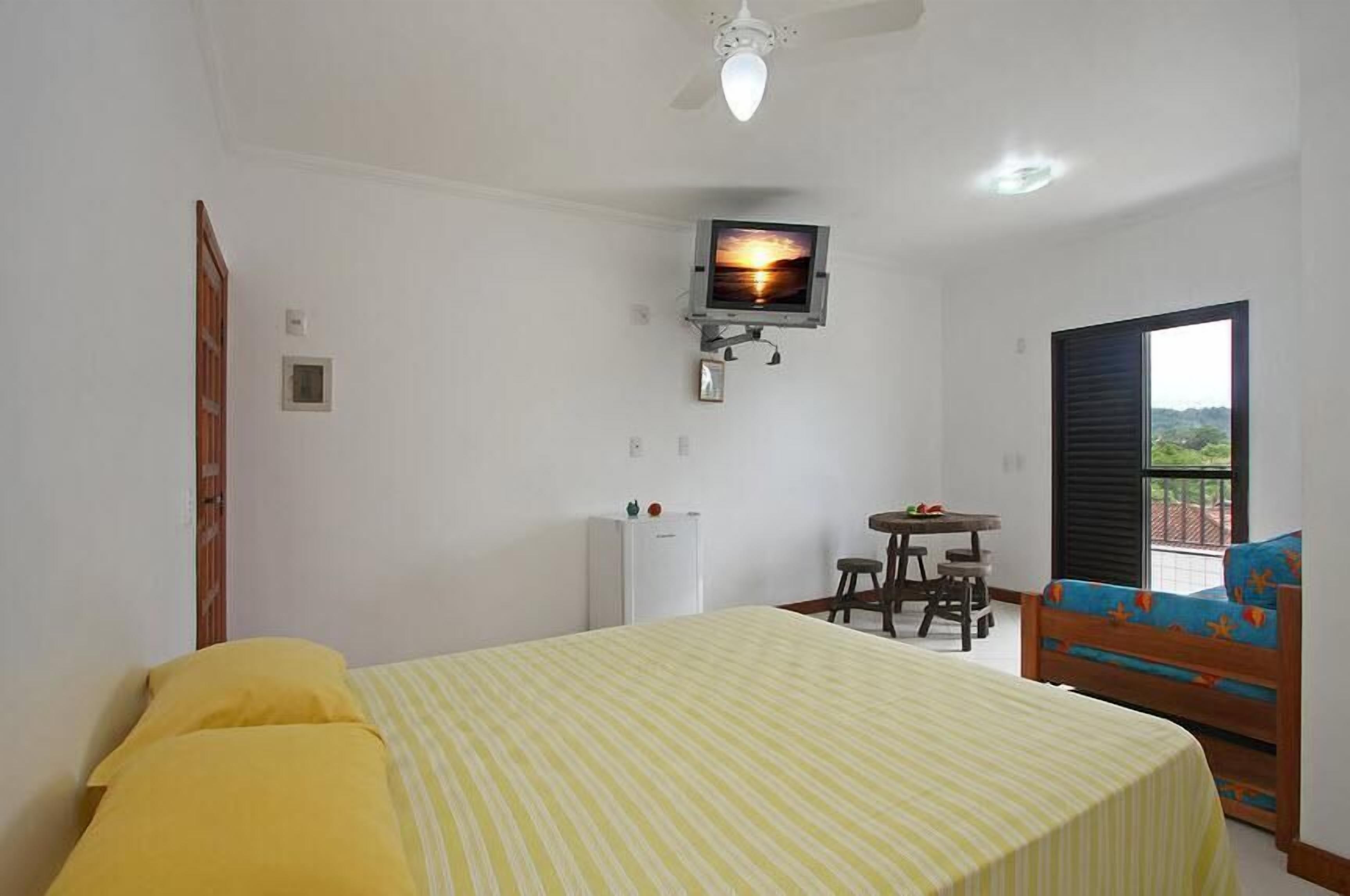 Quarto com uma cama, televisão na parede, mesa pequena com cadeiras e janela com persianas.