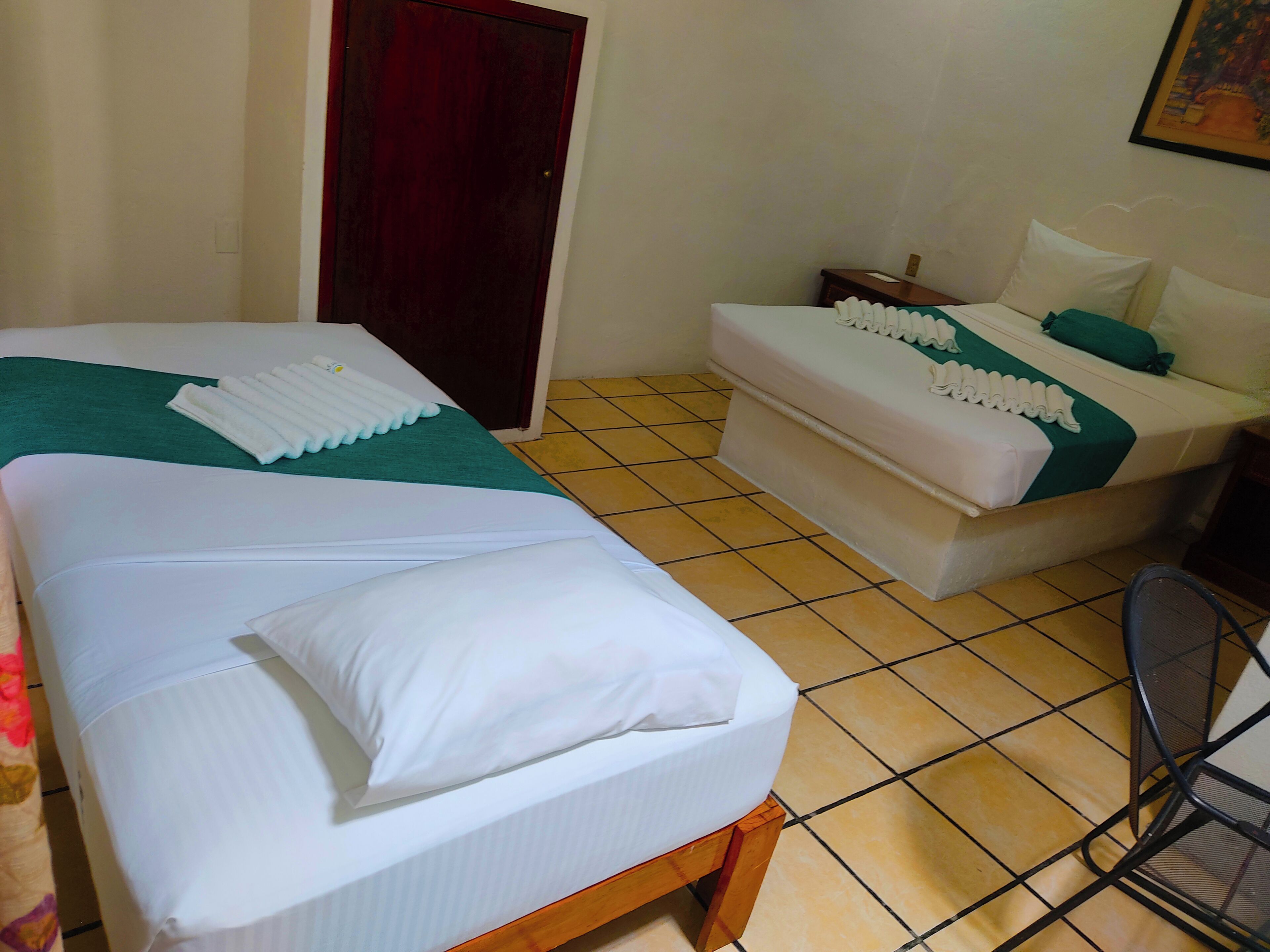 Quarto de hotel com duas camas, piso de madeira, uma porta e uma cadeira.