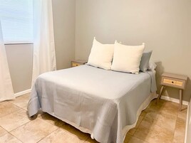 Uma cama bem arrumada com colcha azul clara e almofadas brancas, uma mesa de cabeceira com uma gaveta pequena e uma janela com cortinas brancas.