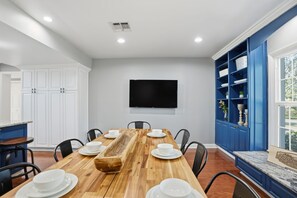 Uma sala de jantar com uma mesa de madeira posta para seis pessoas, cadeiras brancas, um armário azul com objetos decorativos e uma TV de tela plana na parede.