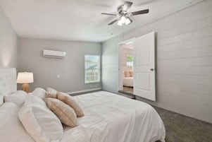 Quarto com cama, ventoinha no teto, aparelho de ar condicionado, janela com persianas e porta que dá para outro cômodo.