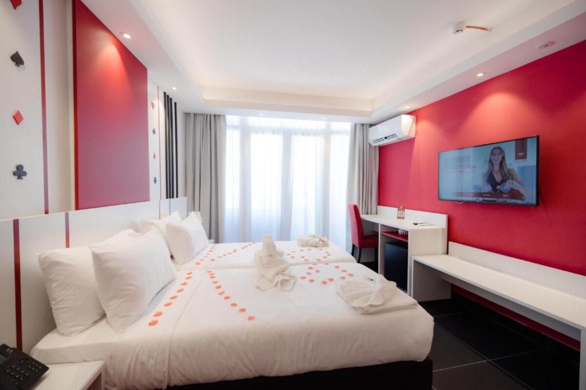 Quarto de hotel com uma cama, uma escrivaninha, uma televisão e uma janela com cortinas.