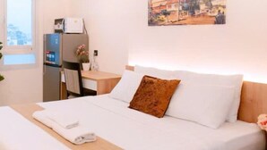 Uma cama bem arrumada com lençóis brancos e um travesseiro marrom, uma mesa de cabeceira com uma luminária e uma pequena escrivaninha com um computador e uma planta.
