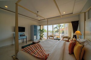 Quarto de hotel com uma cama grande, uma escrivaninha com televisão de tela plana, um espaço de estar com cadeiras e vista para palmeiras e edifícios lá fora.