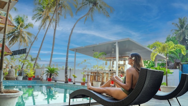 Uma pessoa relaxando em uma cadeira de vime junto a uma piscina, com palmeiras e um espaço coberto ao fundo.