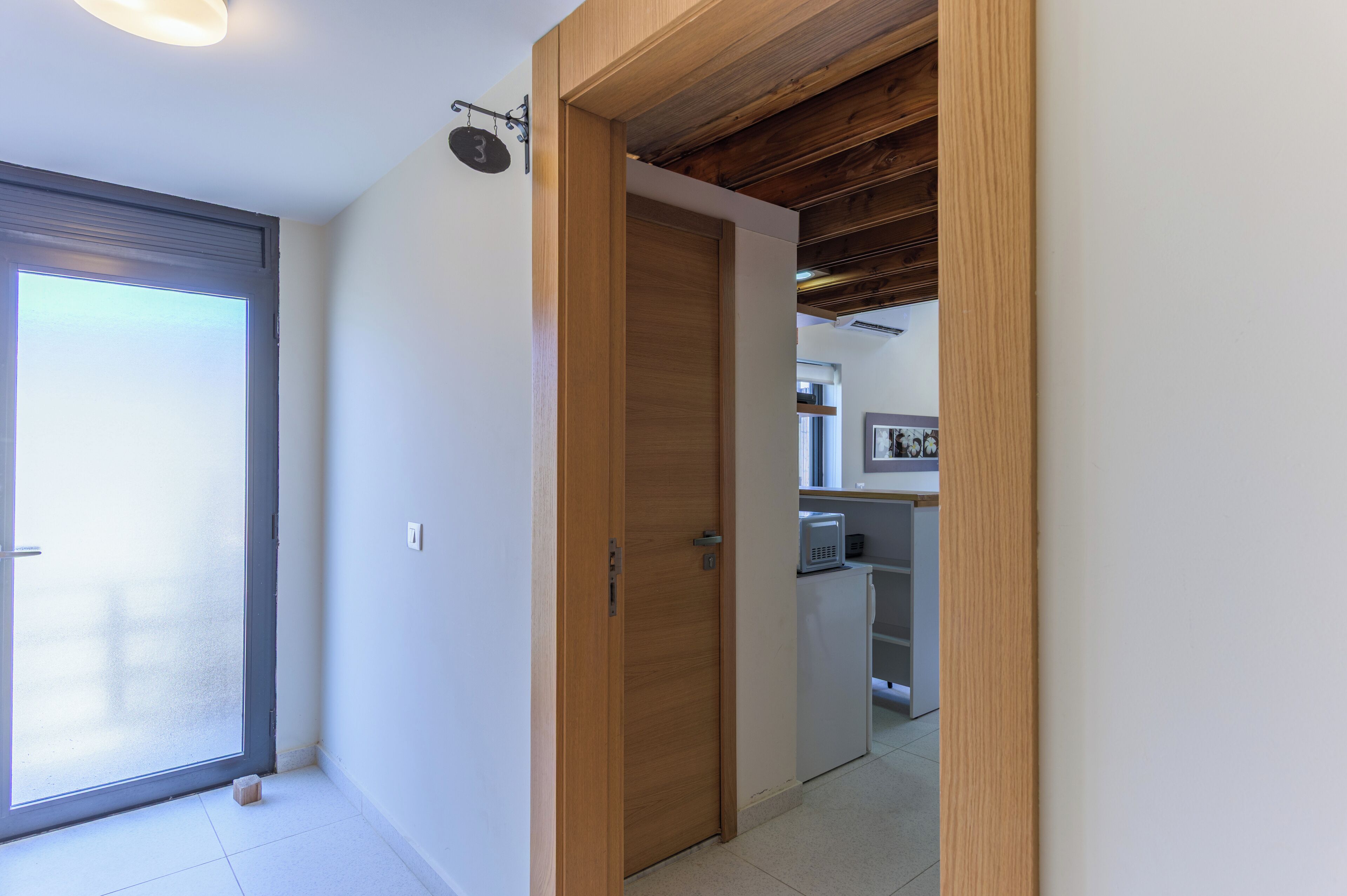 Corredor com porta de madeira, frigorífico branco e área de cozinha com micro-ondas.