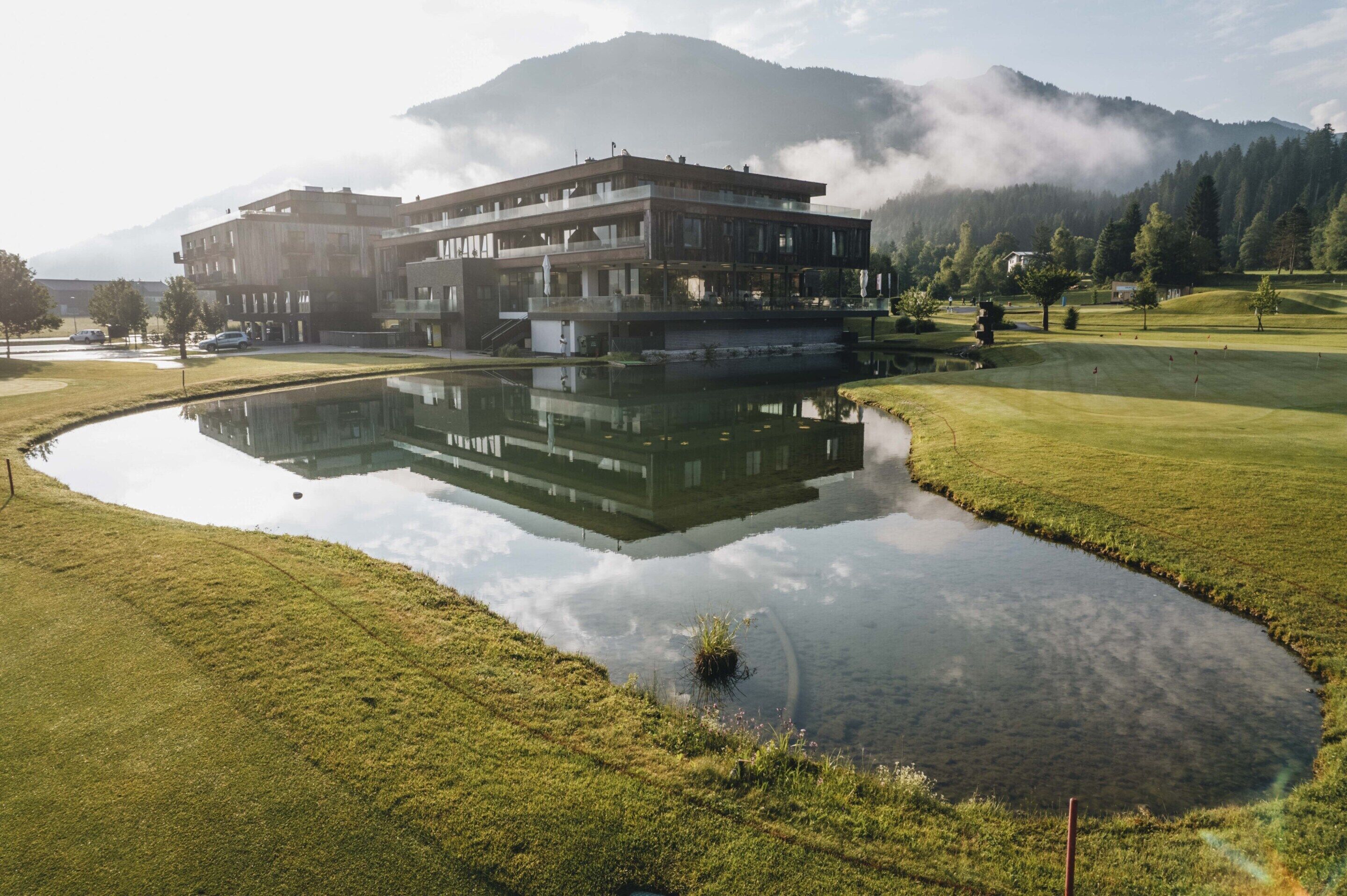 Um edifício moderno refletido em um lago, cercado por um campo de golfe e montanhas.