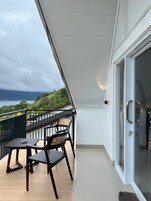 Uma varanda com mesa e cadeiras, uma porta de vidro e vista para o mar e colinas.