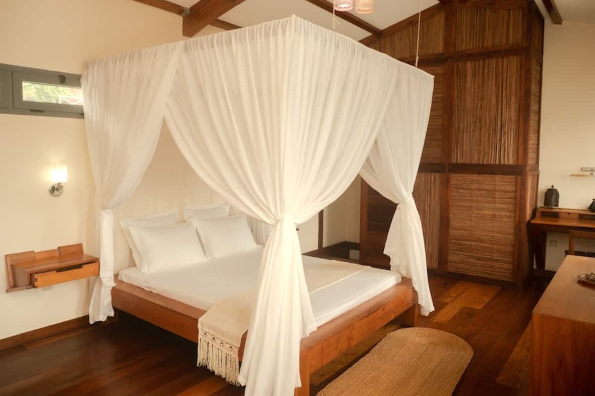 Quarto com cama de dossel, piso de madeira, um banco e uma mesa com abajur.