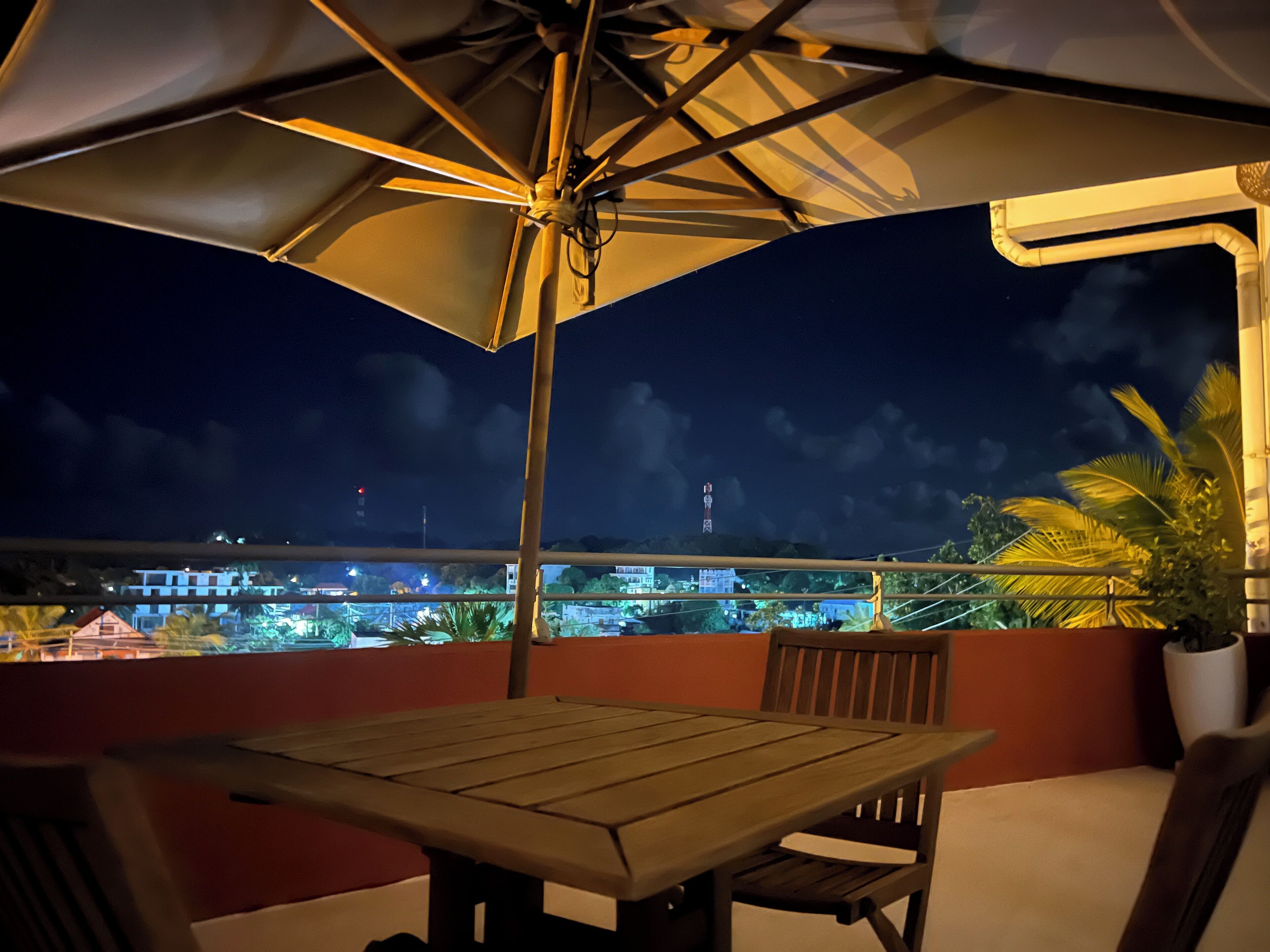Uma varanda com mesa e cadeiras de madeira, um guarda-sol e vista para a paisagem urbana à noite.
