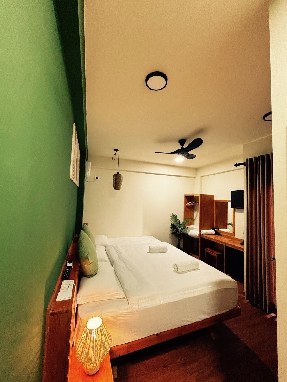 Quarto de hotel com cama, mesa de cabeceira, luminária e ventilador de teto.