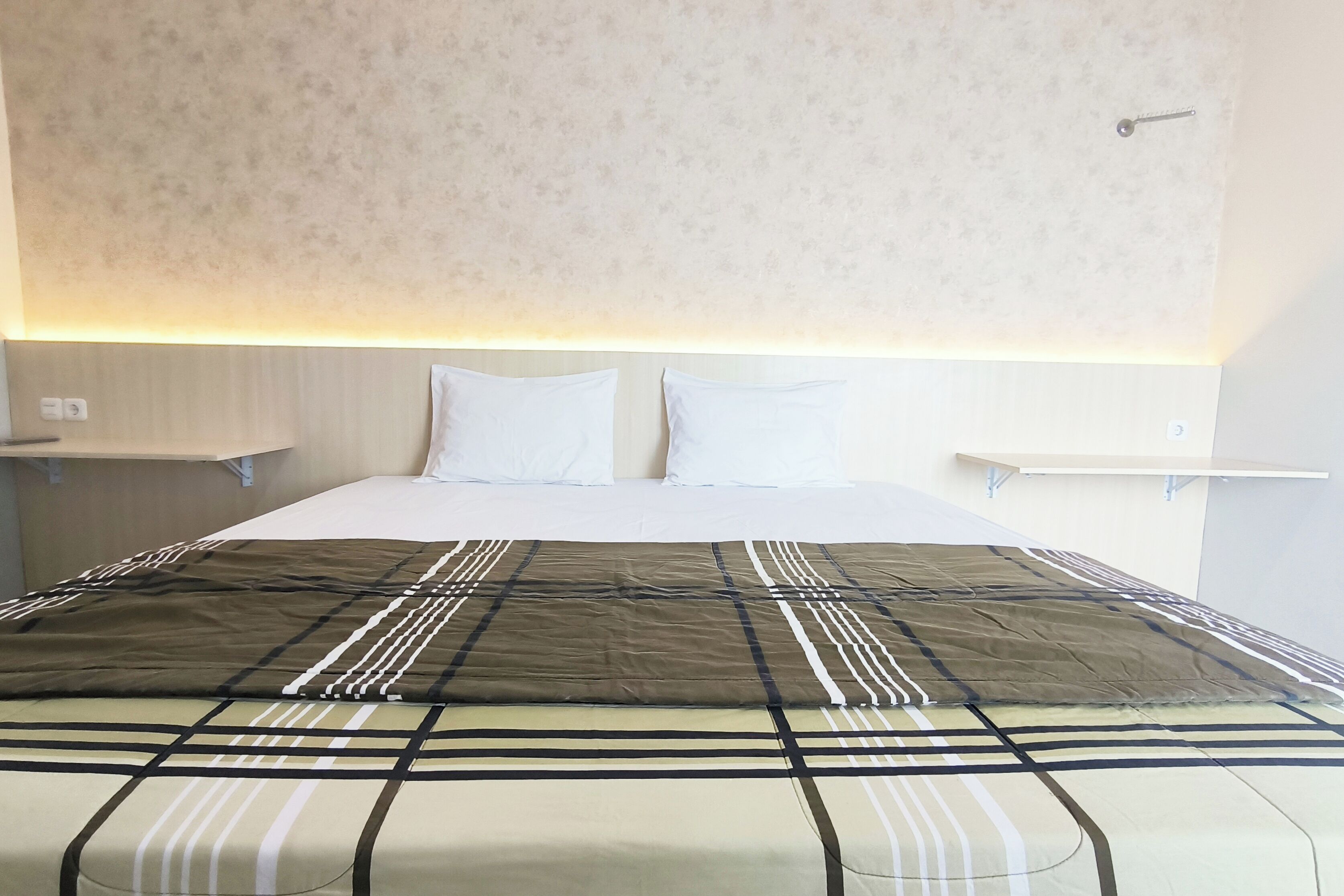 Uma cama bem arrumada com um cobreleito estampado, uma mesa de cabeceira com abajur e um luminário fixado na parede.