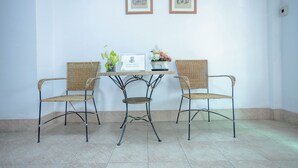 Um quarto com piso de azulejos, uma mesa com duas cadeiras de vime, um certificado emoldurado e um vaso com flores.