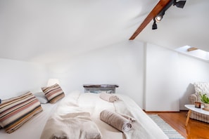 Quarto com teto inclinado, cama com almofadas, mesa de cabeceira com abajur e uma pequena planta.