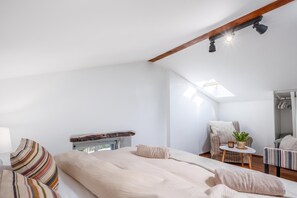Um quarto moderno com teto inclinado, cama com almofadas listradas, uma pequena mesa redonda com uma planta e uma cadeira com almofada listrada.