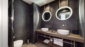 Um banheiro moderno com azulejos escuros, dois espelhos redondos, um aparador de madeira e um vaso sanitário.