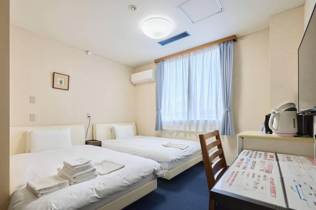 Quarto de hotel com duas camas, uma escrivaninha e chaleira, e uma janela com cortinas.