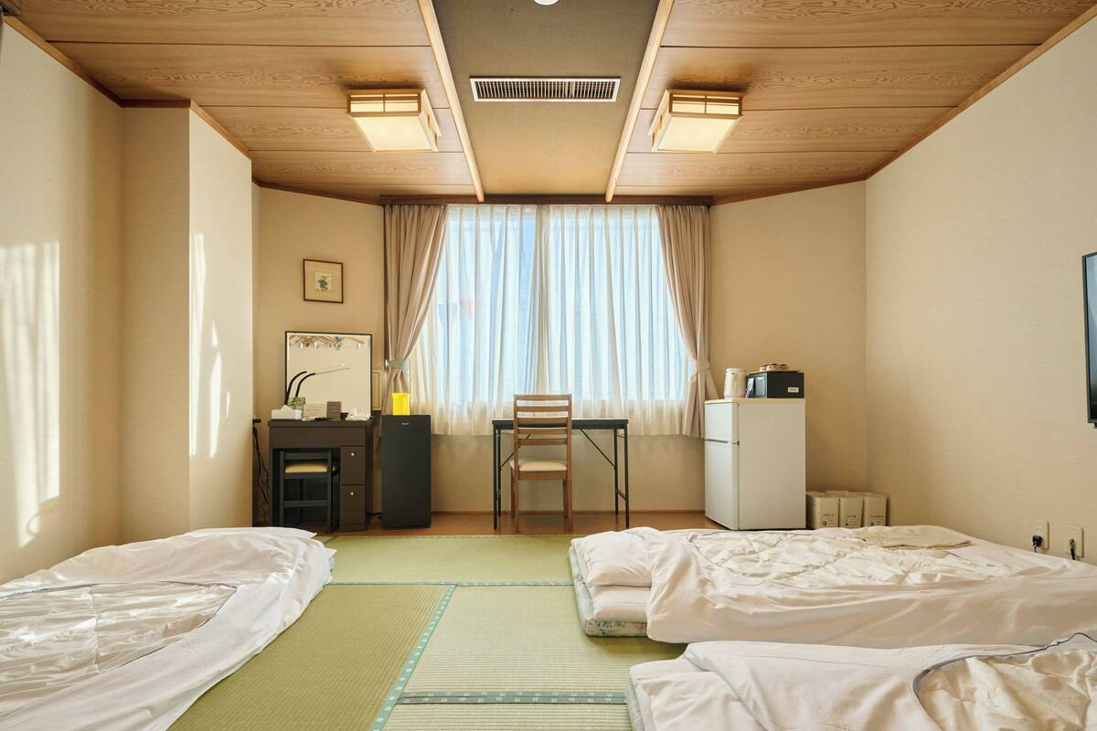 Quarto de hotel com duas camas, uma escrivaninha, uma cadeira, um frigorífico e uma janela com cortinas.