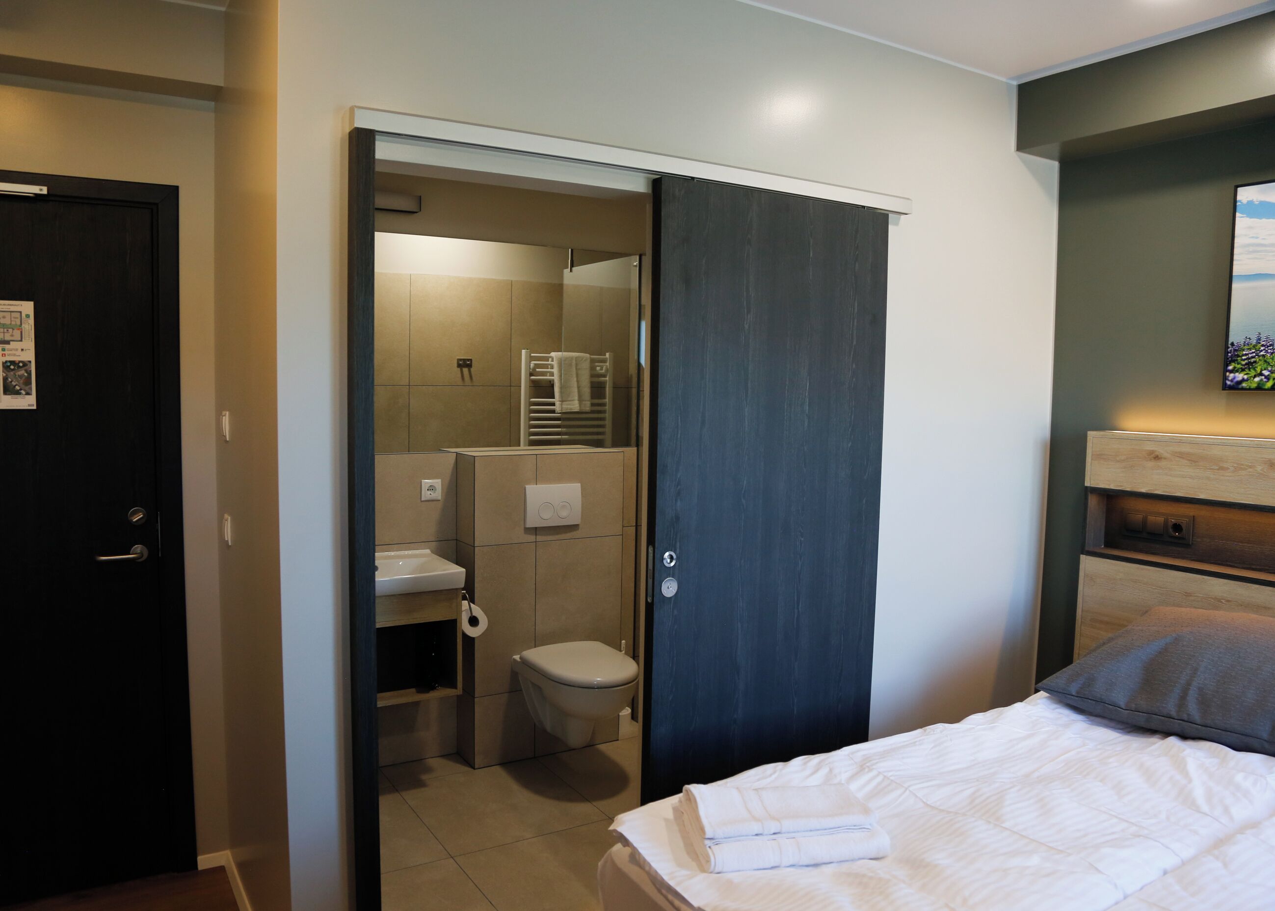 Quarto de hotel com uma cama, banheiro com vaso sanitário e pia, e uma porta escura.