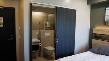 Quarto de hotel com uma cama, banheiro com vaso sanitário e pia, e uma porta escura.