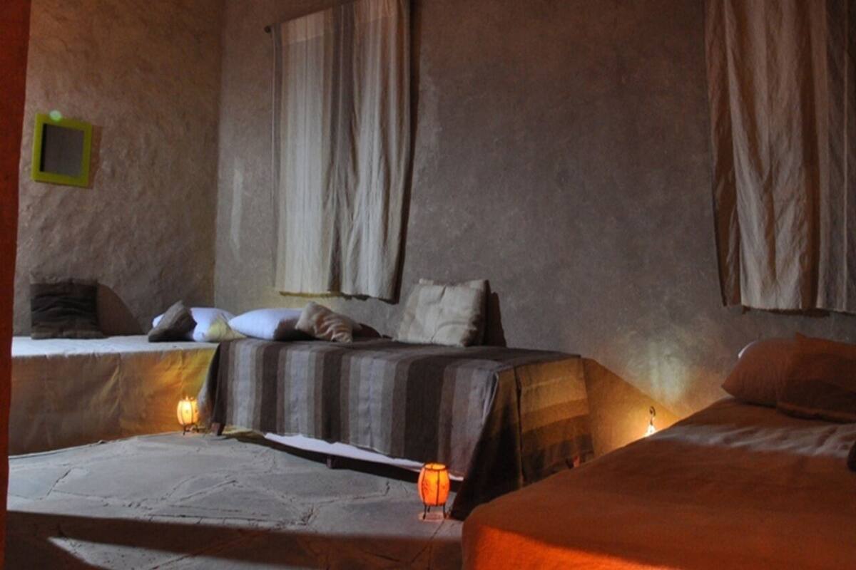 Quarto com duas camas, uma mesinha e prateleira na parede.
