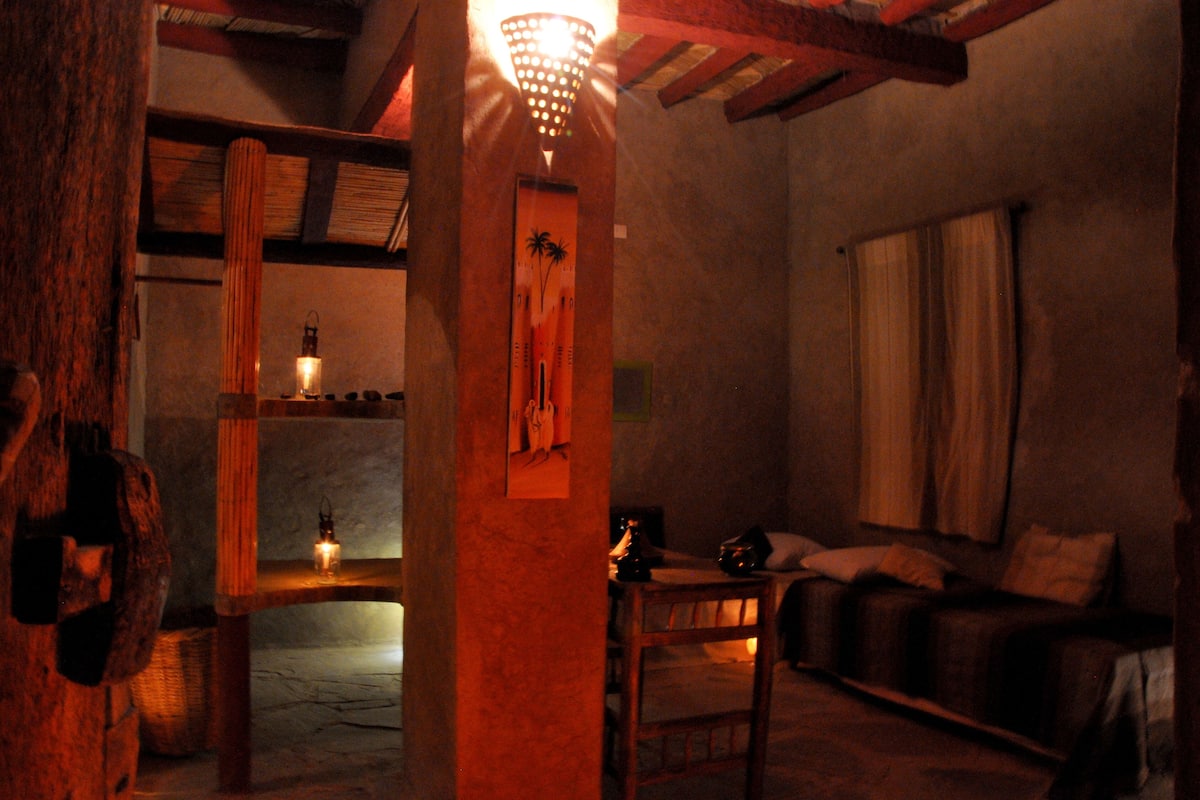 Um quarto com pouca luz, teto de madeira, um sofá, uma mesa e uma estante com livros e uma pequena luminária.