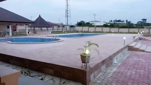 Uma área de piscina com deck revestido, um pequeno jardim com planta em vaso, e um edifício com telhado inclinado ao fundo.