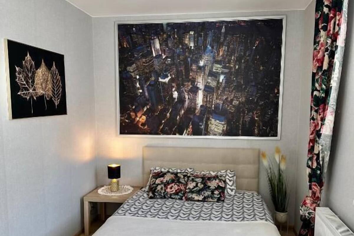 Quarto com uma grande pintura de paisagem urbana, cama com roupa de cama estampada, mesa de cabeceira com abajur e cortina florida.