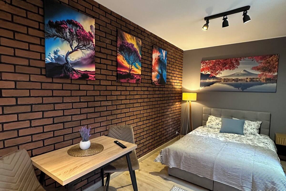 Quarto moderno com parede de tijolos, cama com roupa de cama estampada, mesa de jantar com cadeiras e três quadros.