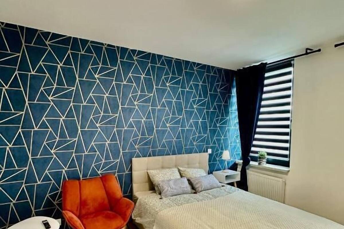 Quarto com parede estampada em padrão geométrico, cama com cabeceira branca, cadeira laranja, mesinha branca e janela com persianas.