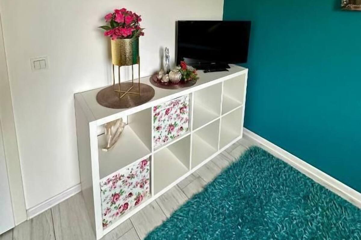 Uma sala de estar com uma parede em tom de verde-água, um console branco com estampas florais, um quadro de uma ponte e uma televisão de tela plana.