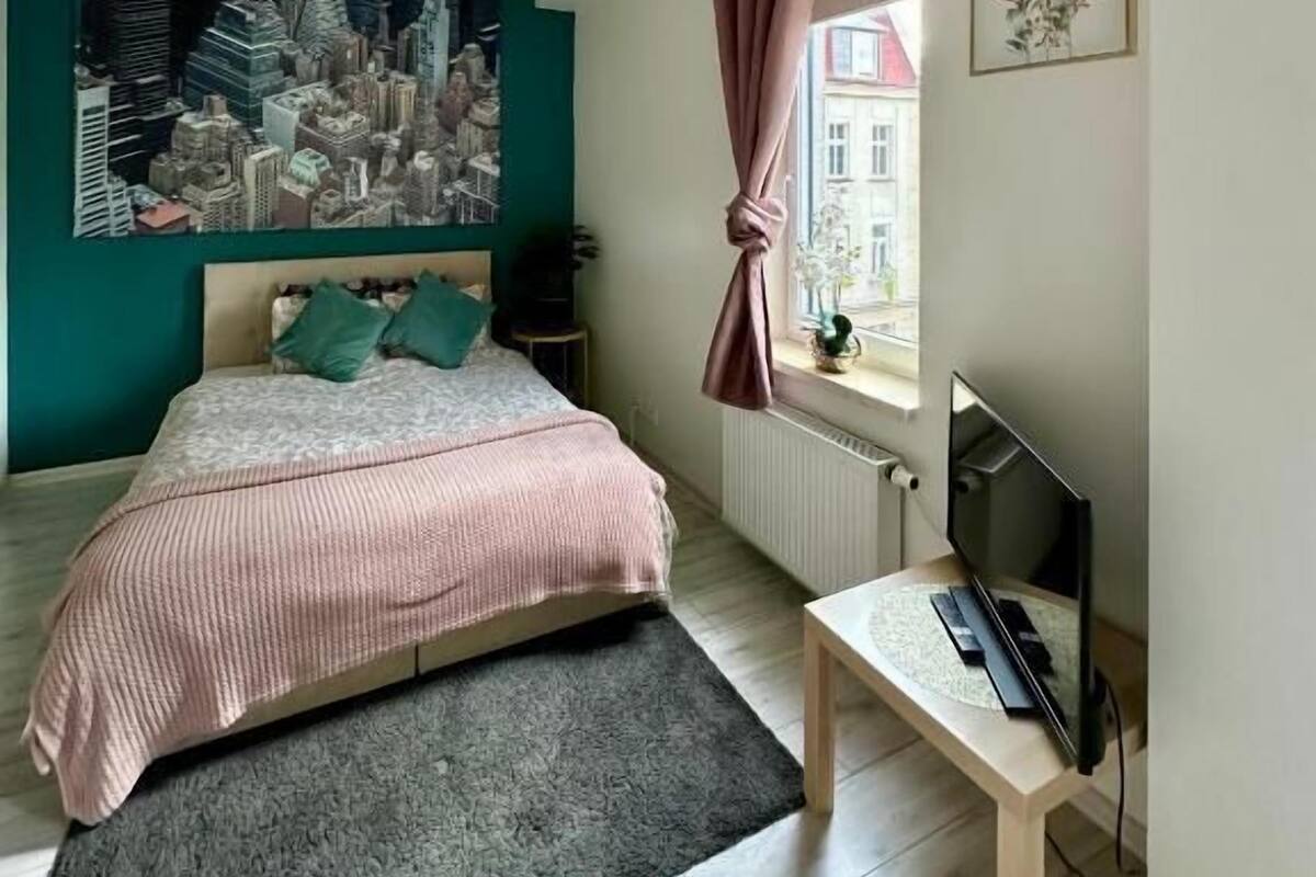 Um quarto com cama, escrivaninha, janela com cortinas, radiador e uma pintura com vista para a cidade.