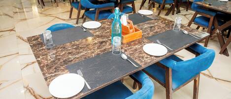 Uma sala de jantar com mesas de tampo em mármore, cadeiras estofadas em azul e louças elegantemente dispostas.