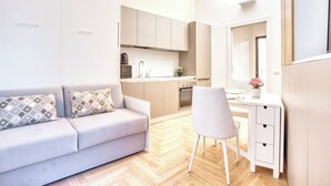 Um apartamento moderno e bem iluminado, com cozinha compacta, sofá com almofadas estampadas, escrivaninha branca e uma cadeira.