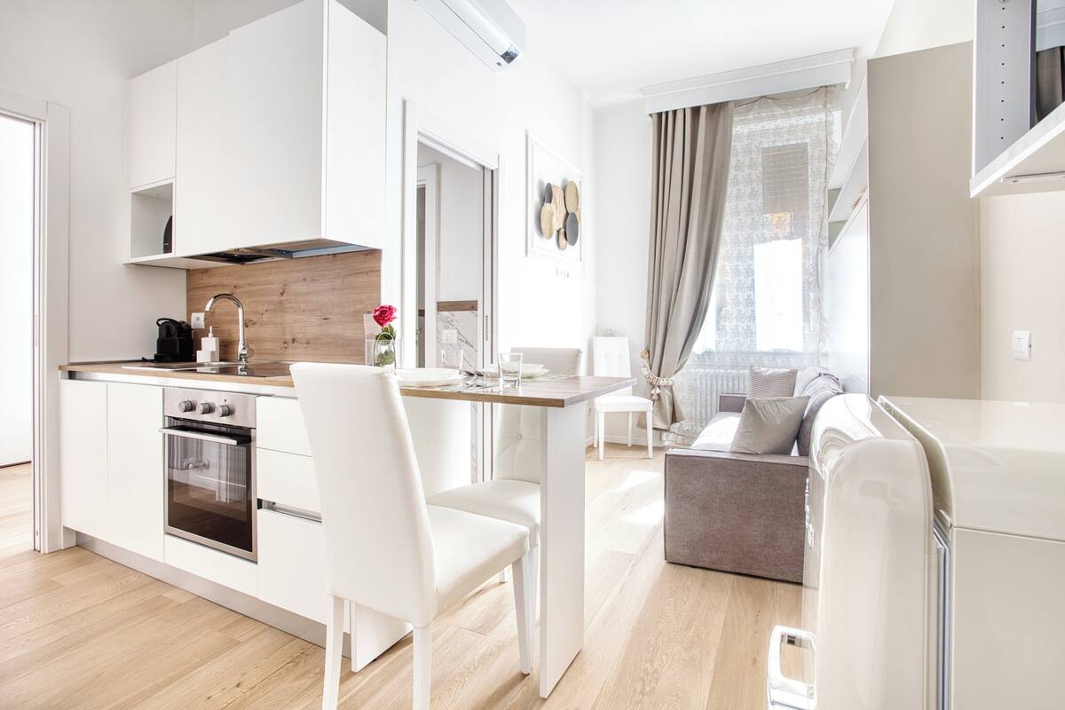 Cozinha moderna com armários brancos, bancada de madeira e forno embutido. Há uma área de jantar com mesa e cadeiras brancas, e um sofá cinza na sala de estar.