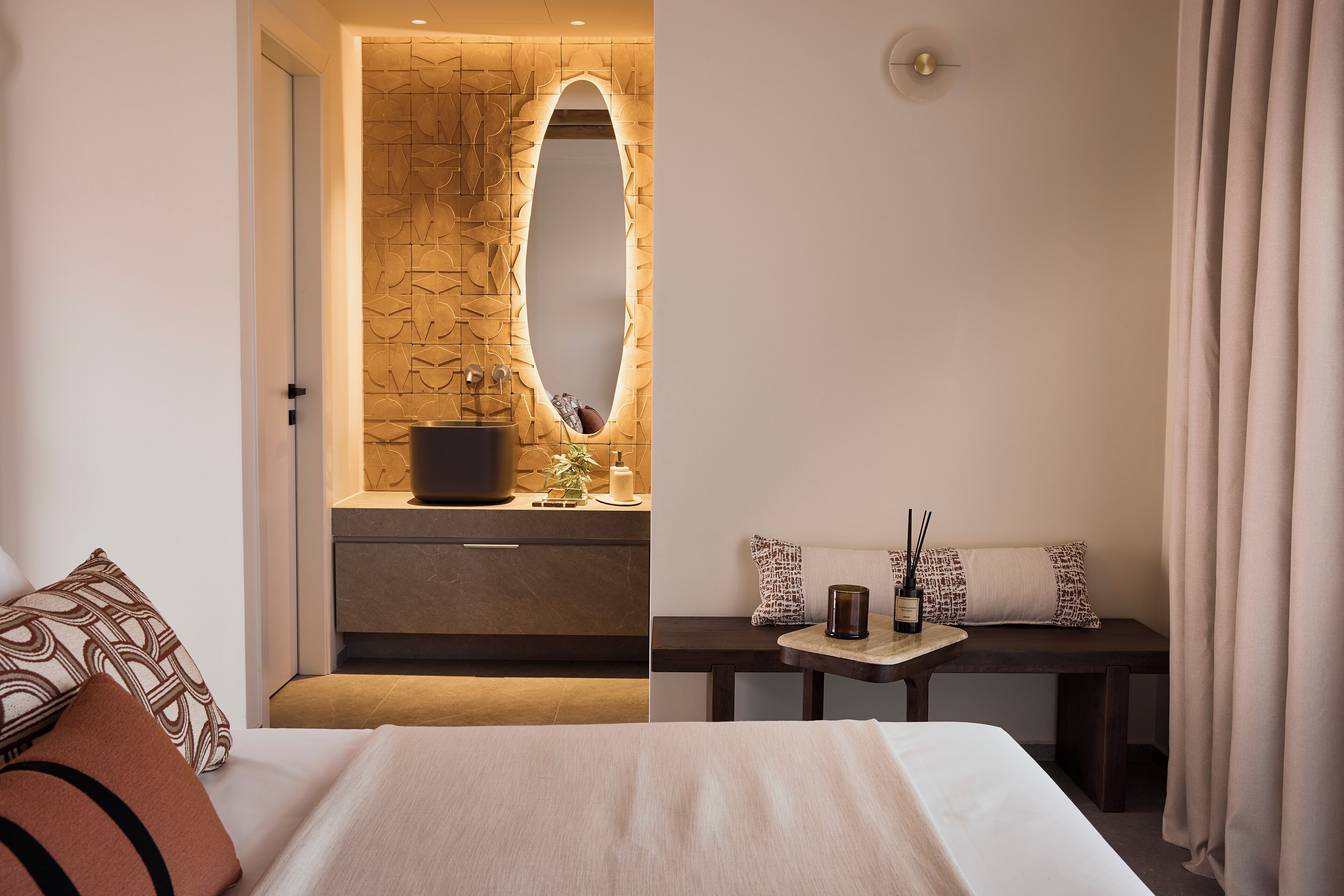 Quarto de hotel com cama, mesa de cabeceira com abajur, espelho e banheiro com lavatório e suporte para toalhas.