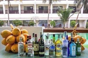 Uma mesa com várias garrafas de bebidas e um cacho de cocos em frente a um edifício com várias varandas e palmeiras.