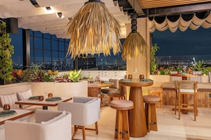 Um bar no terraço com vista para a cidade à noite, apresentando uma luminária pendente grande, áreas de estar e uma variedade de plantas.