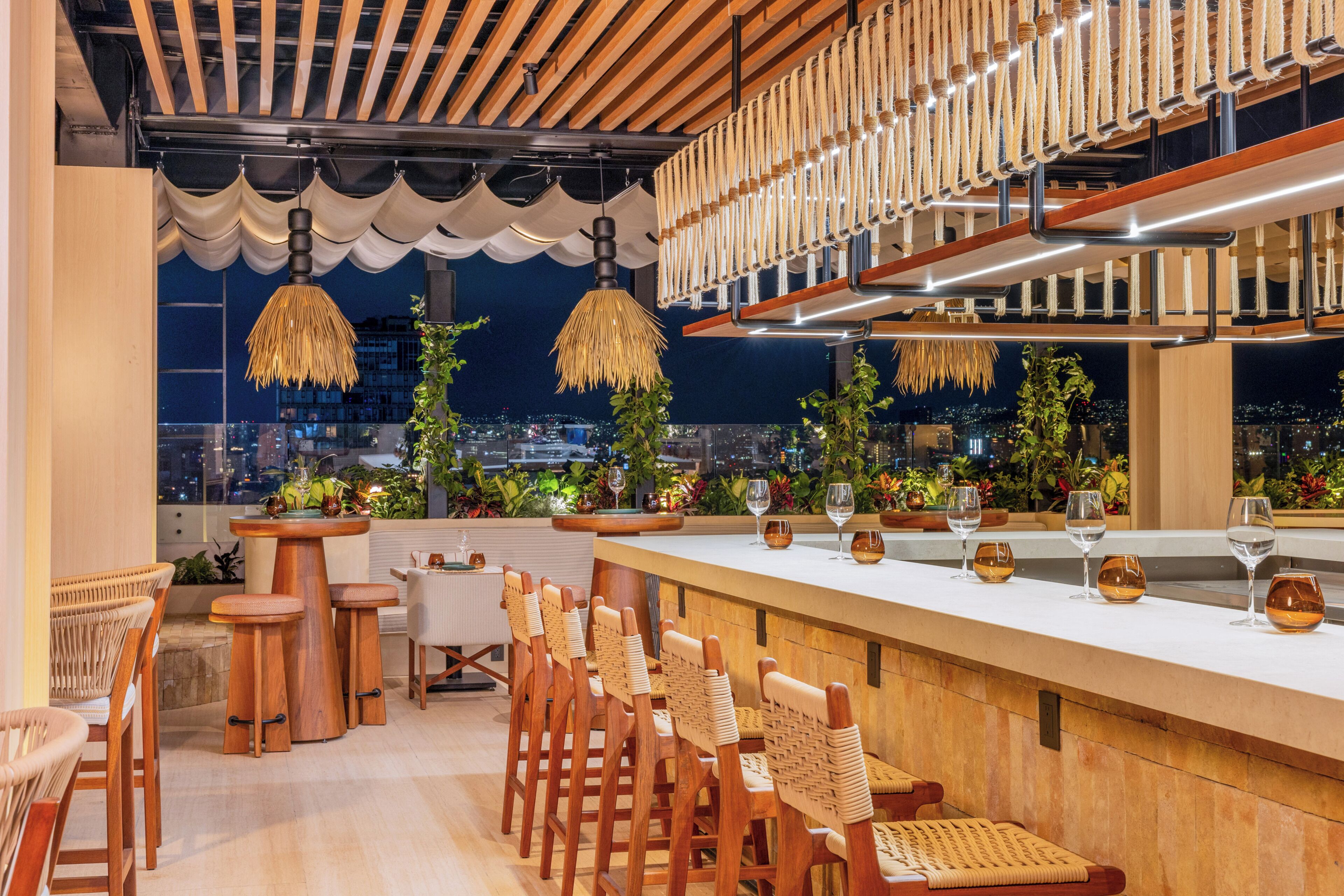 Um bar no terraço com mobiliário de madeira, luminárias pendentes e vista para a cidade.